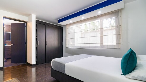 Quarto moderno com cama, janela com persianas, guarda-roupa e porta para outro cômodo.