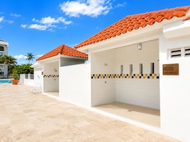 Uma área junto à piscina com piso de azulejos, telhado de telhas vermelhas e um edifício branco com parede revestida.