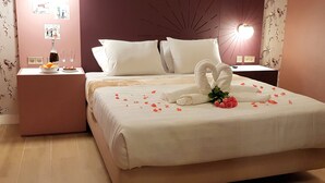 Cama bem arrumada com arranjo de toalhas em formato de coração, mesa de cabeceira com garrafa e copos, e parede com quadros emoldurados.
