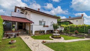 Uma casa de dois andares com um caminho de pedra que leva à entrada, um pequeno jardim com uma fonte e um telhado de telhas vermelhas.