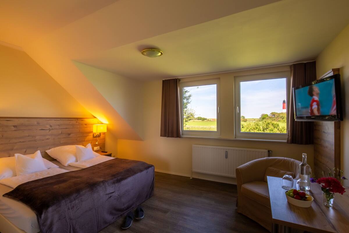 Quarto de hotel com uma cama, televisão, janela com vista para o verde, mesa com uma garrafa e flores, e um radiador.