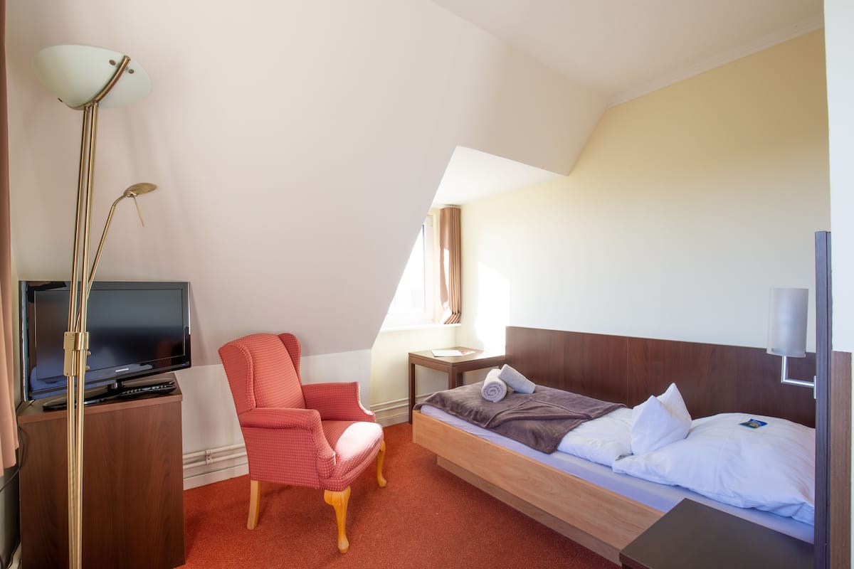 Quarto de hotel com uma cama, uma escrivaninha com luminária, uma televisão e uma cadeira vermelha.