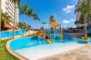 Uma piscina com escorregas e área de recreação aquática, cercada por palmeiras e um prédio com restaurante de pizza e churrascaria.