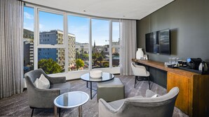 Quarto de hotel moderno com uma janela ampla, televisão de tela plana, um pequeno bar e área de estar.
