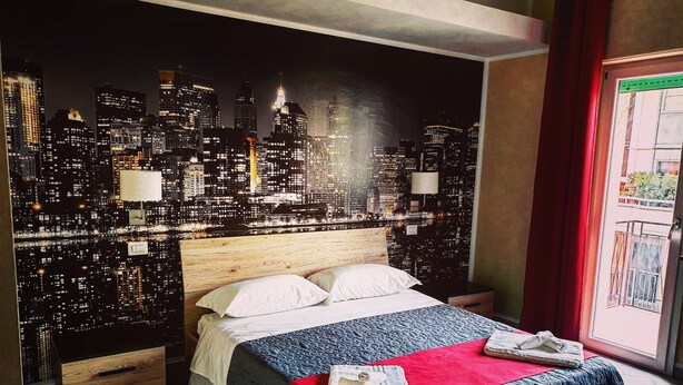 Quarto de hotel com mural da paisagem urbana na parede, cabeceira de madeira, cama com colcha azul e vermelha, e mesa de cabeceira com abajur.