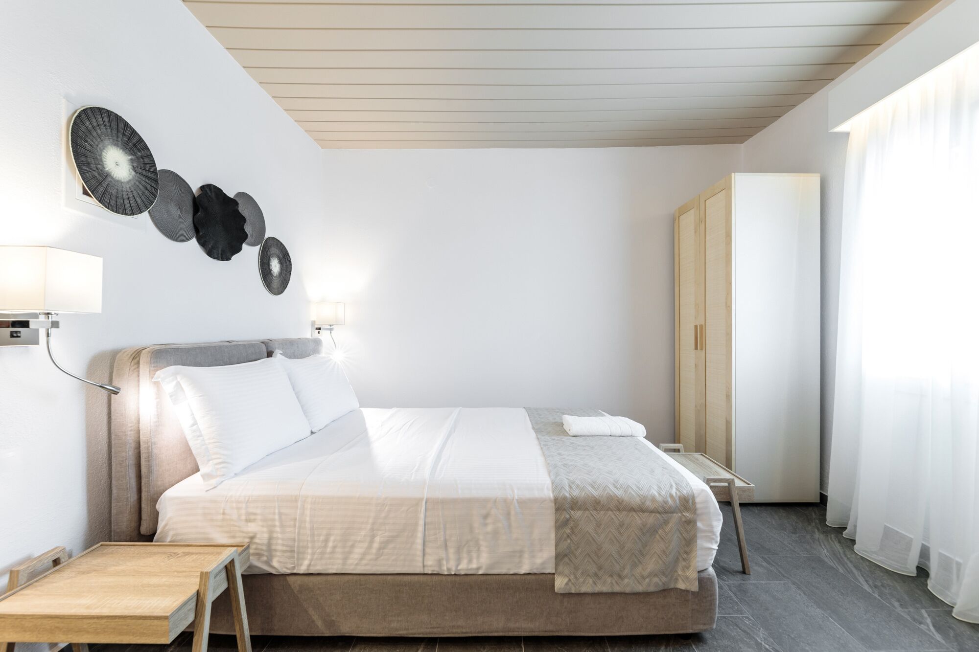 Quarto de hotel moderno com uma cama, mesas de cabeceira, guarda-roupa e vista para o exterior.