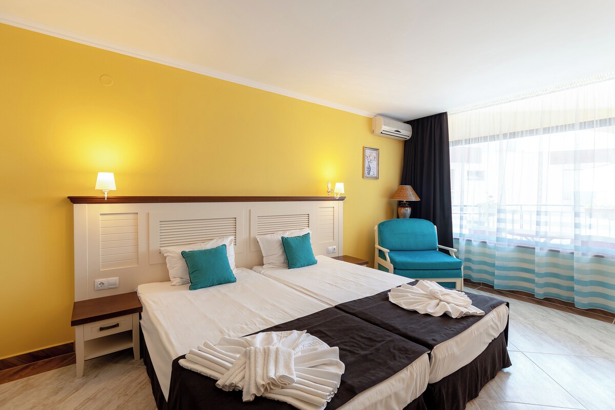 Quarto de hotel com duas camas, uma cadeira e uma janela com cortinas.