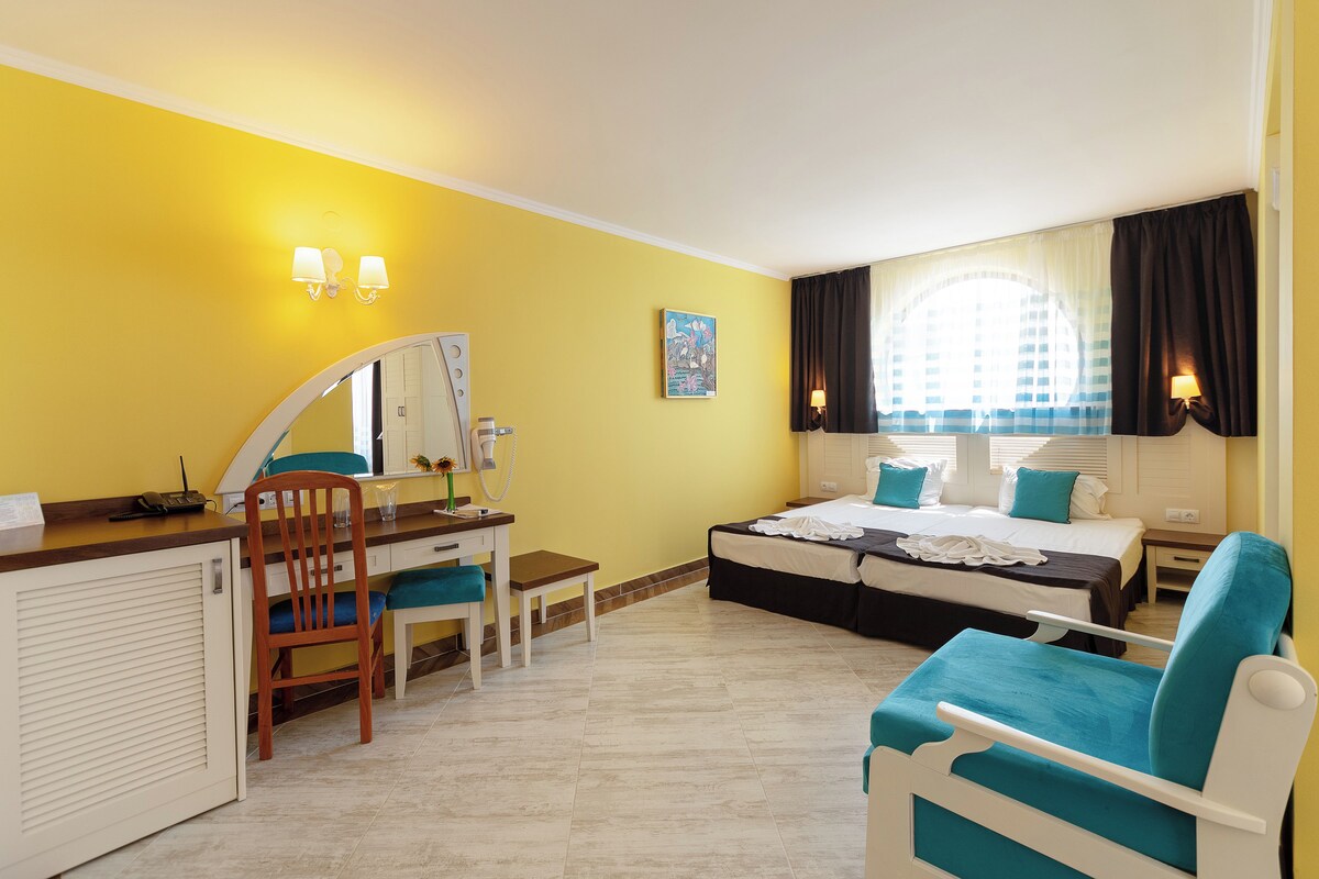 Quarto de hotel com uma cama, uma escrivaninha com cadeira, um espelho, uma janela com cortinas e um quadro na parede.
