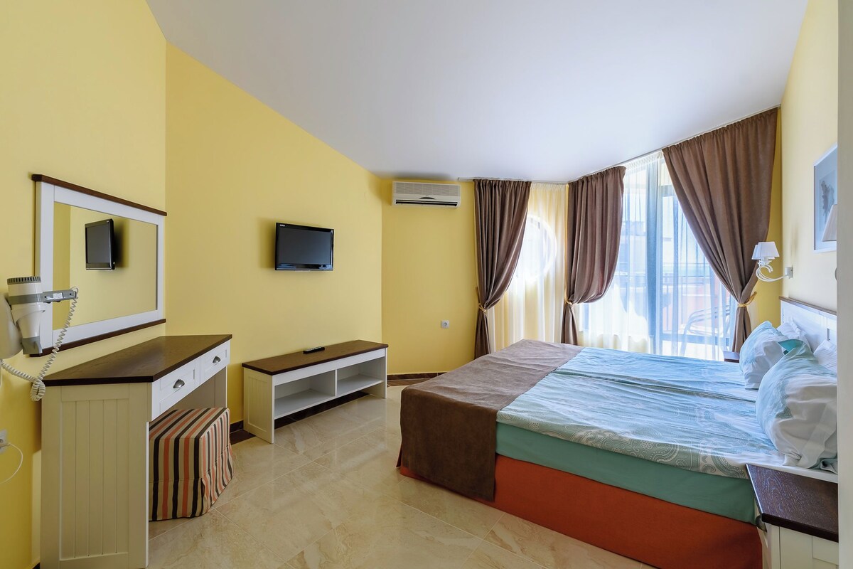 Quarto de hotel com cama, televisão, escrivaninha com telefone, espelho e janela com cortinas.