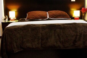 Cama bem arrumada com colcha marrom escura e dois travesseiros, com duas mesas de cabeceira com luminárias e um pequeno vaso de flores.