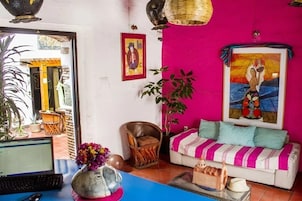 Uma sala de estar vibrante com uma parede rosa, um sofá branco com almofadas verde-água, uma cadeira de vime e um laptop sobre uma mesa azul.