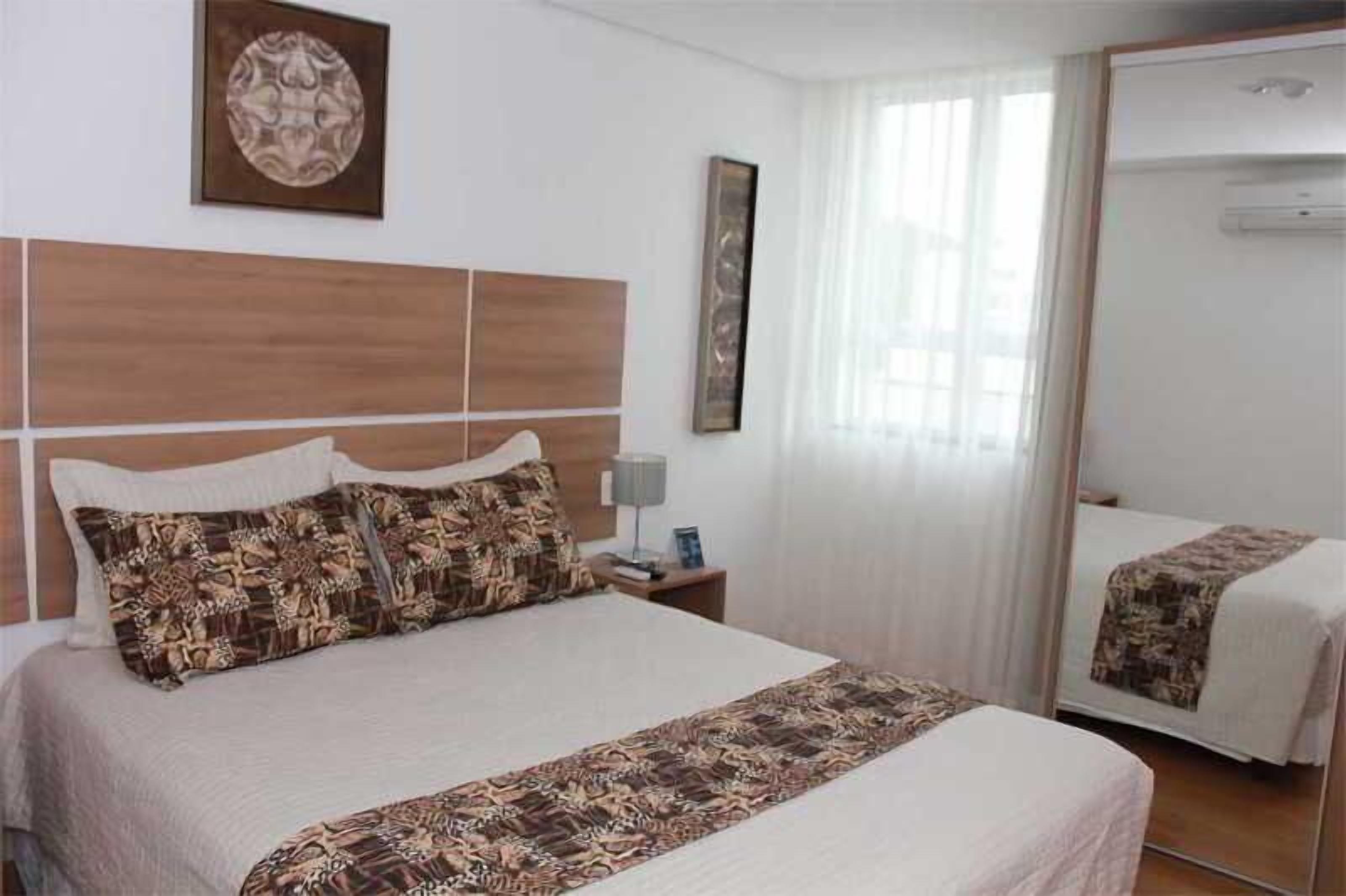 Quarto de hotel com uma cama grande, mesa de cabeceira, espelho e janela com cortinas.