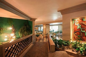 Corredor de hotel com parede verde, corrimão de madeira e vista para a área de refeições.