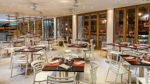 Restaurante com várias mesas, cadeiras, ventiladores de teto e janelas amplas.