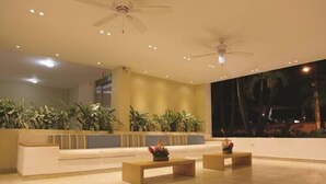 Um hall amplo com teto baixo, iluminação embutida e duas ventoinhas no teto. Há plantas em vasos ao longo das paredes e um balcão de recepção.