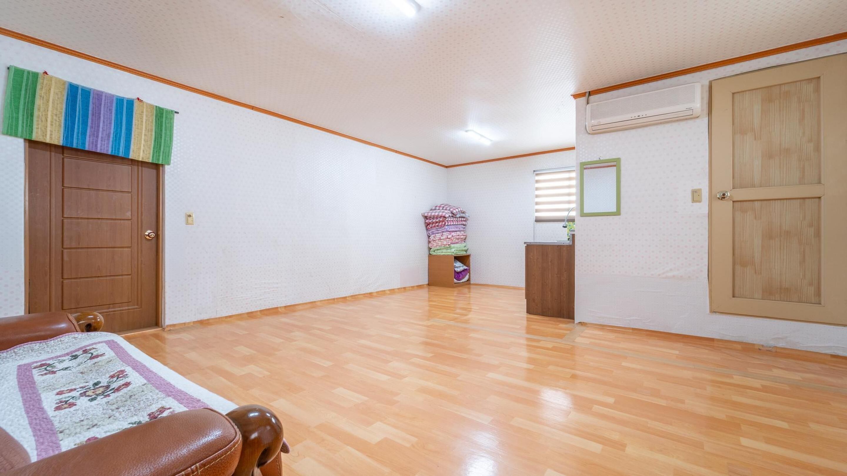 Um quarto espaçoso com piso de madeira, uma porta marrom, uma pequena cozinha compacta e uma janela com persianas.