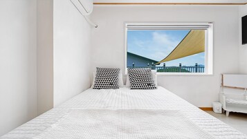 Quarto com cama, dois travesseiros estampados, janela com vista e mesa de cabeceira branca.