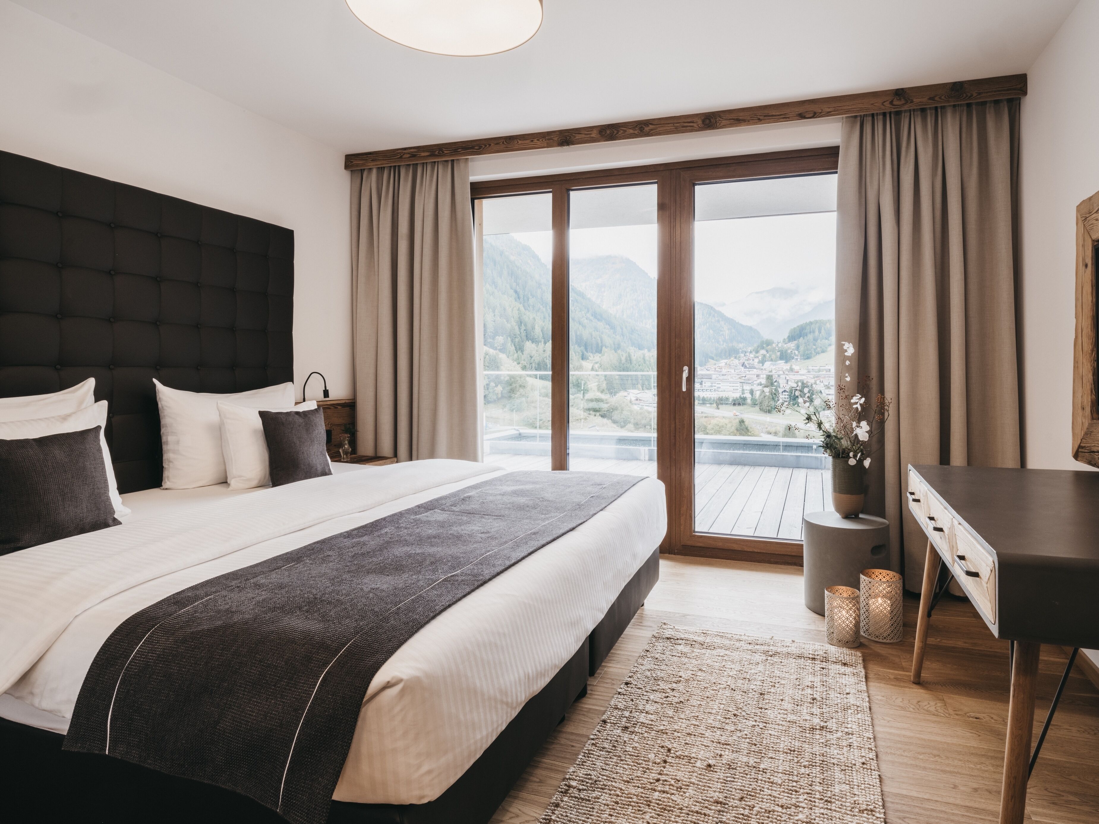 Quarto moderno com uma cama grande, cabeceira de madeira, mesa de cabeceira, cômoda e vista para montanhas através de uma porta de vidro deslizante.