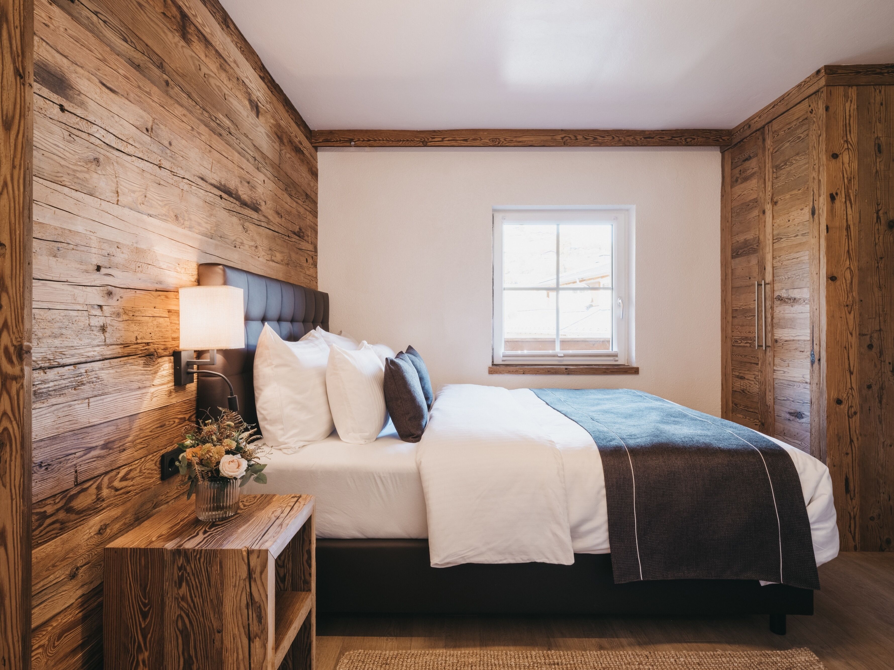 Quarto com cabeceira de madeira, mesa de cabeceira com abajur, janela e cama com cobertor.