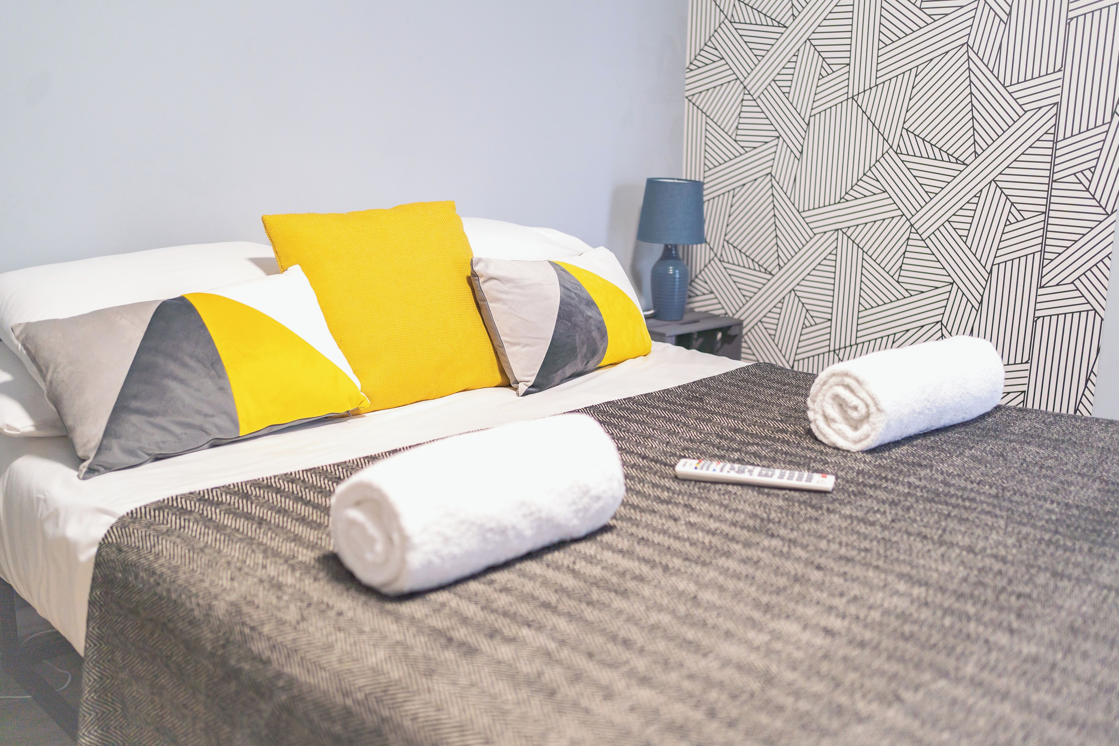 Uma cama bem arrumada com um edredom cinza e branco, dois travesseiros em amarelo e cinza, uma toalha branca enrolada e um controle remoto sobre a cama.