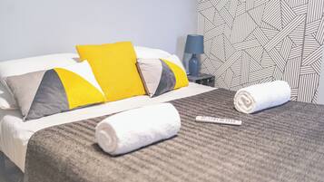 Uma cama bem arrumada com um edredom cinza e branco, dois travesseiros em amarelo e cinza, uma toalha branca enrolada e um controle remoto sobre a cama.