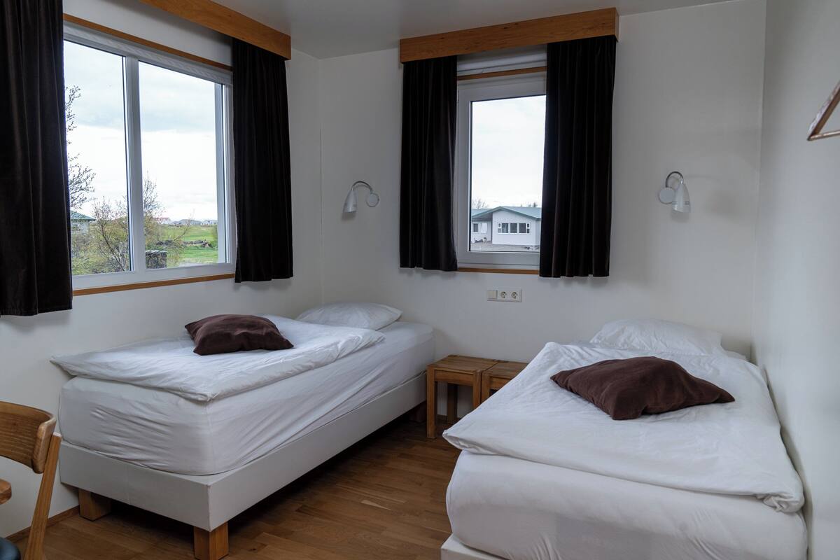 Quarto com duas camas de solteiro, uma cadeira de madeira, uma mesinha e uma janela com cortinas.