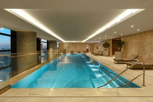 Piscina interior moderna com barreira de vidro, espreguiçadeiras e vista para a cidade.