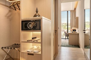 Quarto de hotel com cofre embutido, sapateira e vista da cidade através de amplas janelas.