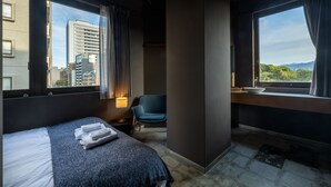 Quarto de hotel com cama, uma cadeira, escrivaninha com luminária e vista para a paisagem urbana e montanhas.