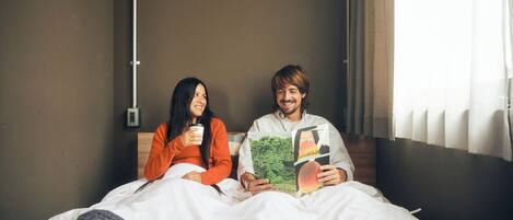 Um casal sentado em uma cama com um livro e uma xícara, em um quarto com parede lisa e uma janela com cortinas.