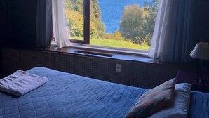 Quarto com cama grande, janela com cortinas e vista para um lago e montanhas.