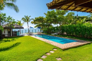 Uma piscina com um deck ao redor, um pequeno edifício e vegetação exuberante.