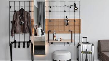 Quarto de hotel moderno com painel quadriculado, cabide suspenso, espelho, prateleira pequena com uma câmera e uma garrafa, mala com adesivo 'Eats Sleep Fly' e um banquinho redondo.