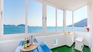 Quarto com grandes janelas que oferecem vista para o mar e colinas ao longe. Há uma pequena mesa com uma garrafa, um copo e uma vela.