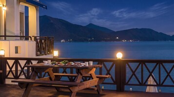 Um deck de madeira com mesa para piquenique montada para refeições, com vista para um corpo d'água calmo e montanhas ao entardecer.