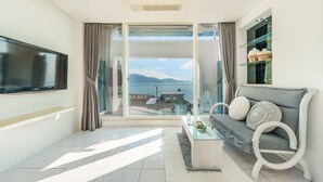 Uma sala de estar moderna com uma televisão grande, um sofá com almofadas decorativas, uma mesa de centro de vidro e vista para o mar através de amplas janelas.