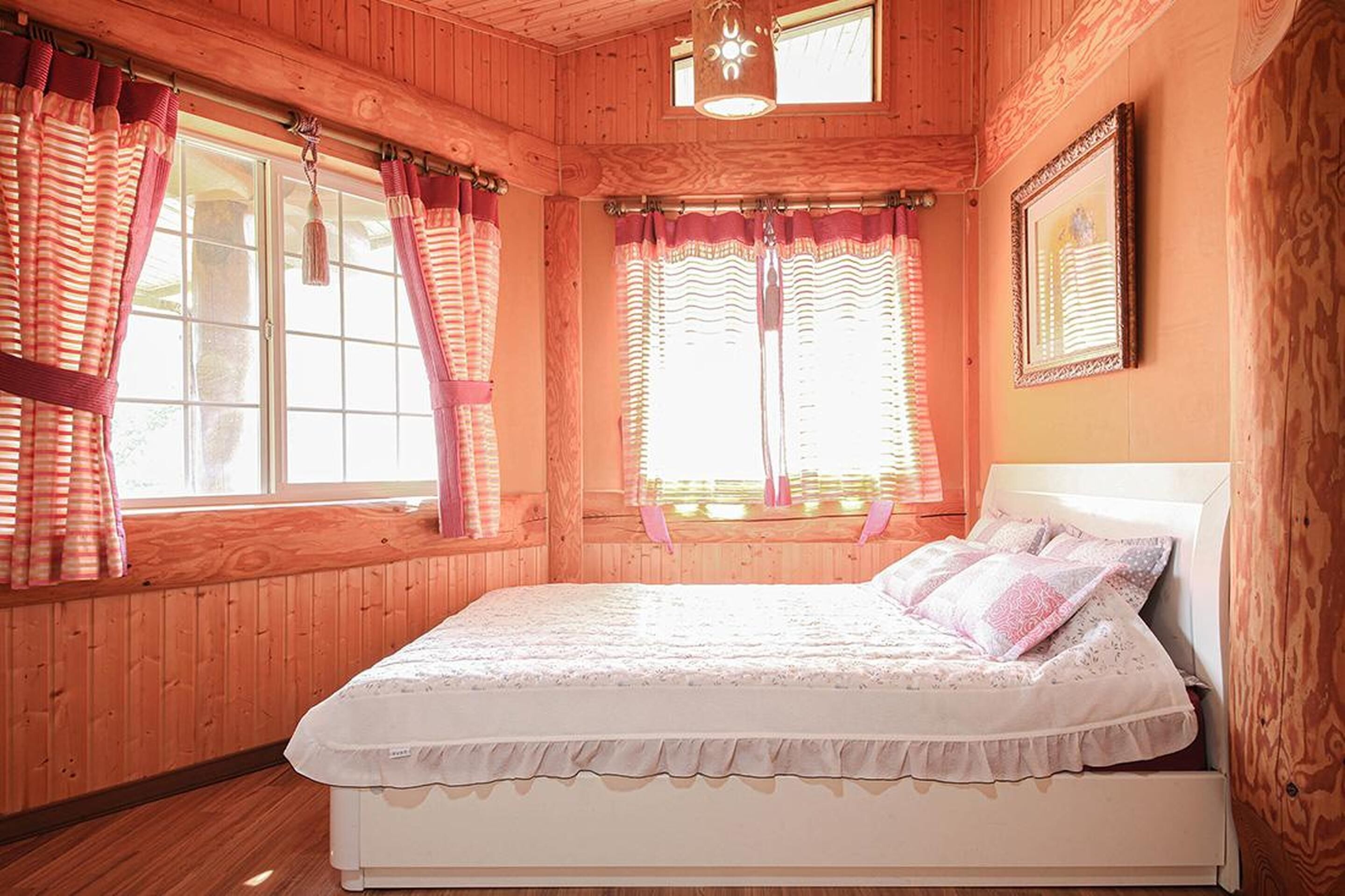 Quarto com painéis de madeira, uma cama, duas janelas com cortinas e um quadro na parede.