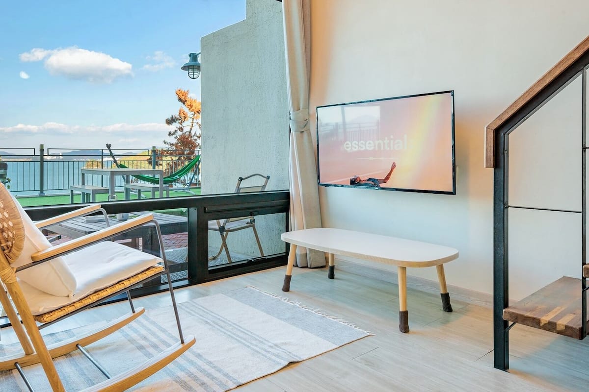 Uma varanda com vista para um jardim, uma cadeira de balanço de madeira, uma mesa pequena e uma televisão fixada na parede.