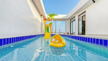 Uma piscina interior com chão de azulejos azuis, um flutuador amarelo em formato de argola e um escorrega com decoração de palmeira.