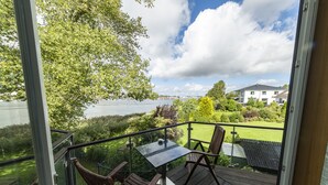 Varanda com mesa e cadeiras, com vista para um lago e um jardim bem cuidado, com uma casa ao fundo.