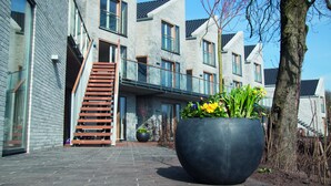 Um edifício moderno com uma escadaria que leva a uma varanda, um grande vaso com flores e uma árvore em primeiro plano.