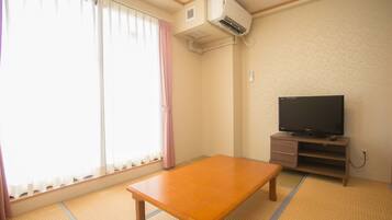Um quarto com piso de tatame, uma porta deslizante com cortinas, uma televisão sobre um suporte de madeira e um aparelho de ar condicionado fixado na parede.