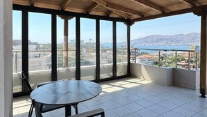 Área externa coberta com mesa e cadeiras, grandes janelas com vista para a paisagem urbana e montanhas.