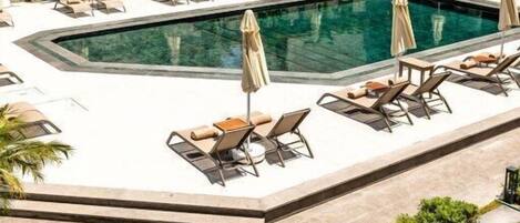Piscina com espreguiçadeiras e guarda-sóis, rodeada por vegetação e edifícios.