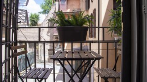 Varanda com planta em vaso, bancos de madeira e vista para um edifício com varandas e plantas.
