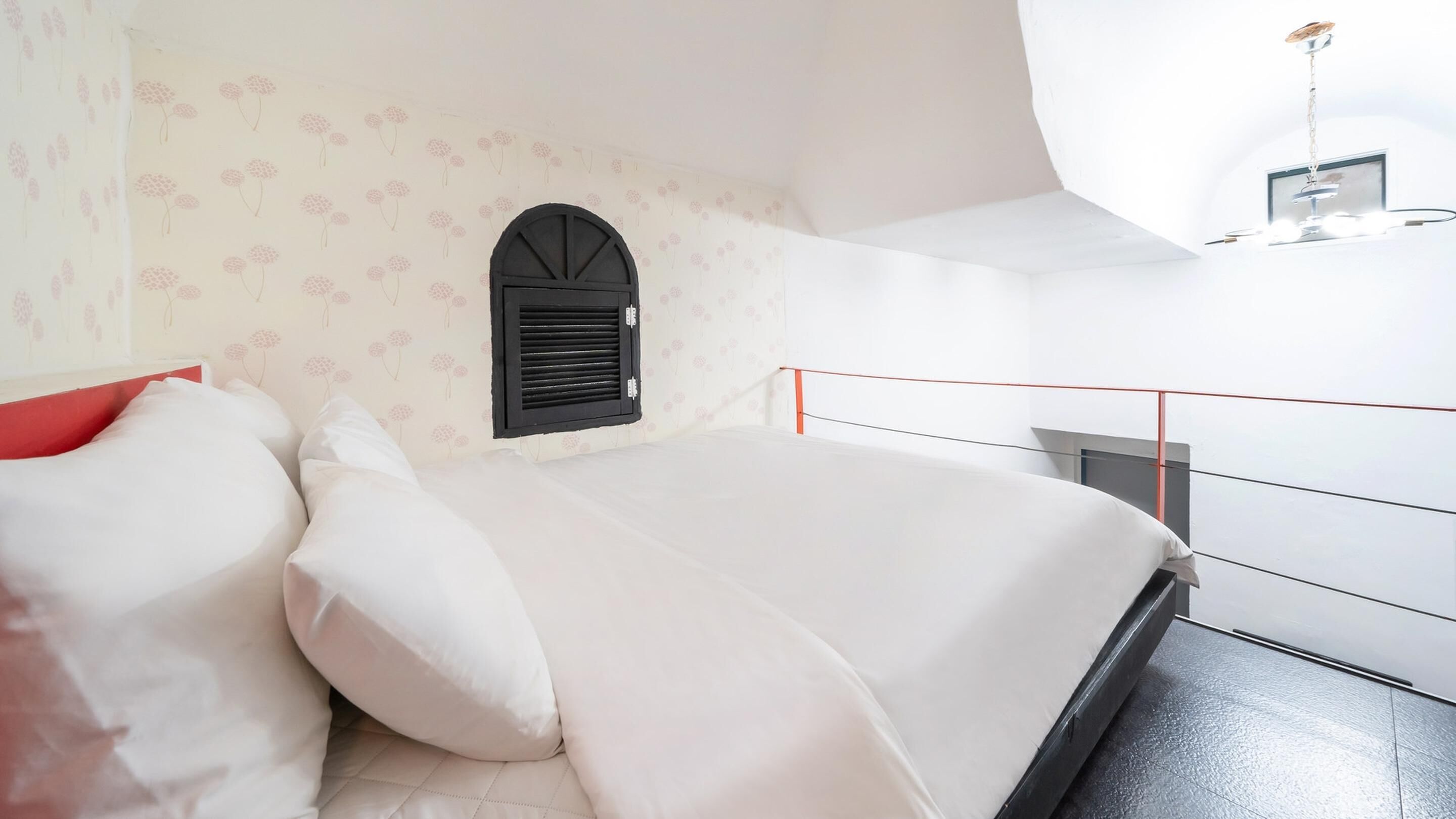 Quarto moderno com cama, pequena janela com persianas, corrimão vermelho e luminária de teto.