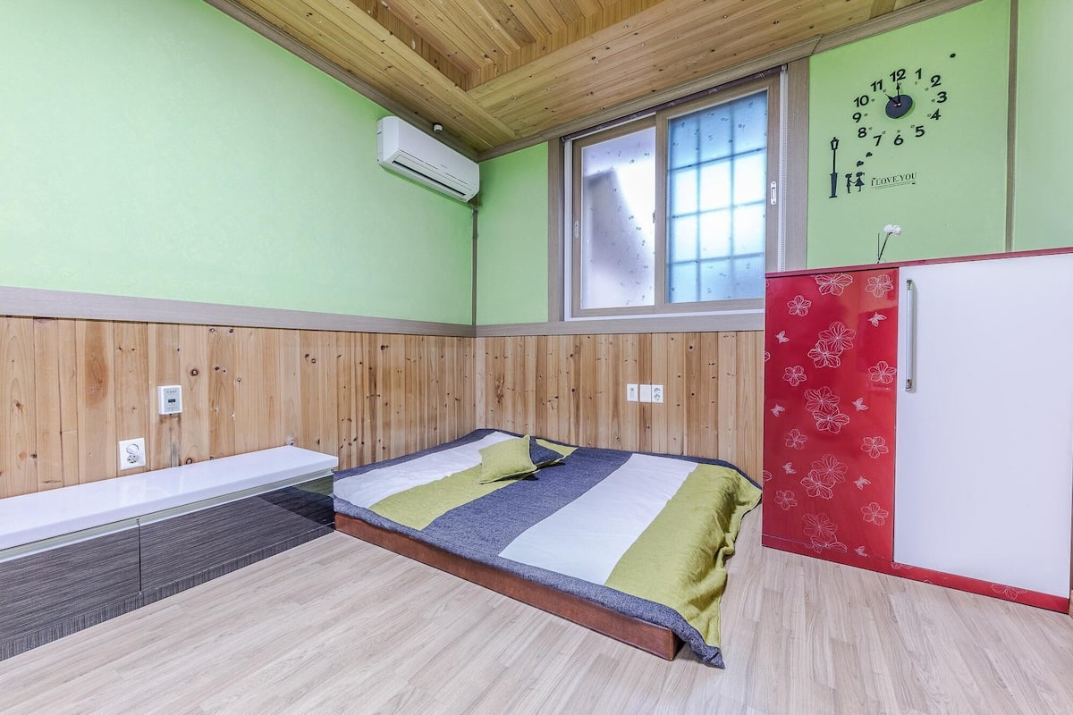 Um quarto com uma cama, uma janela, um relógio de parede e uma geladeira vermelha.
