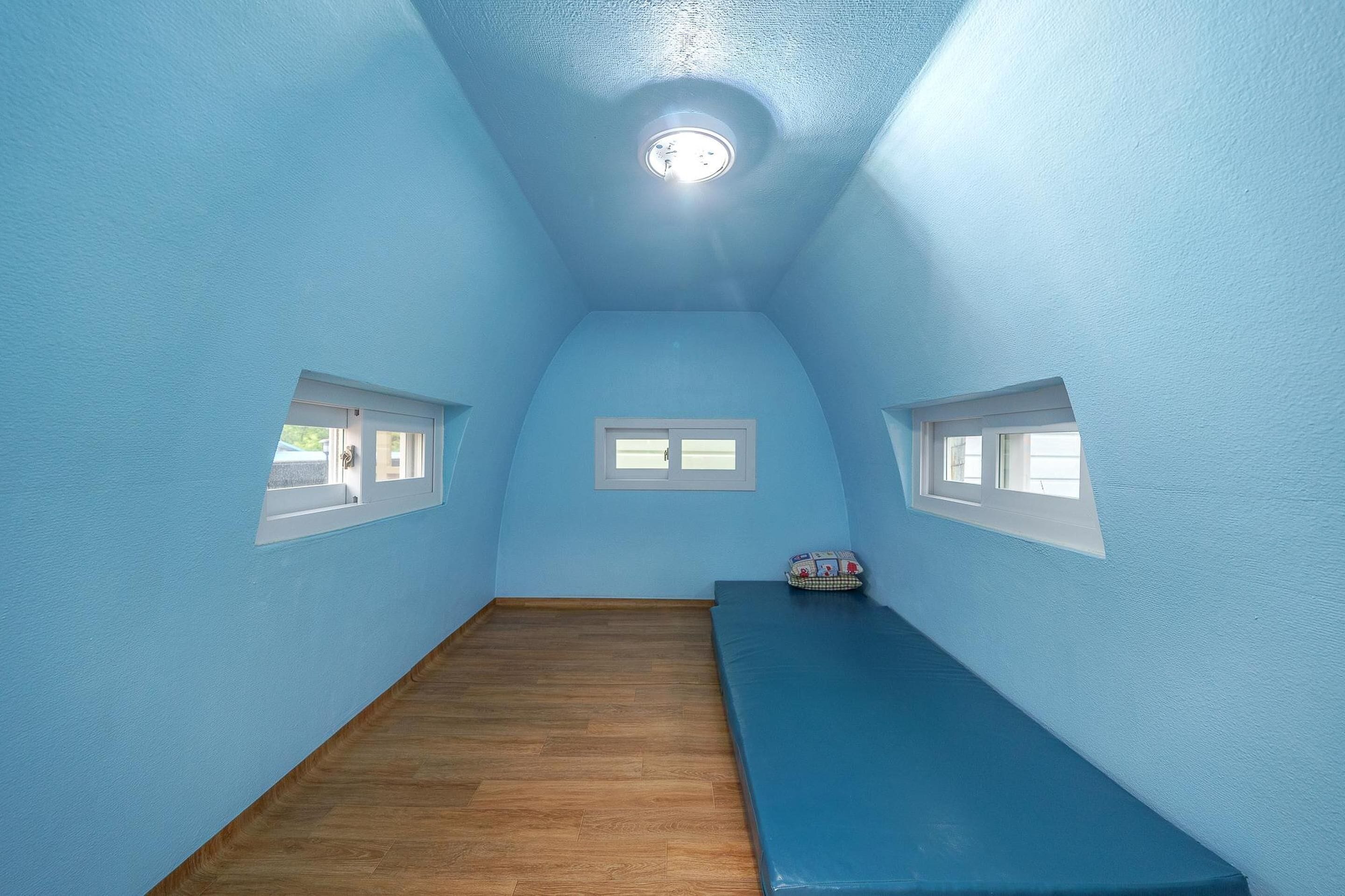 Quarto com paredes azuis, chão de madeira, uma cama beliche e duas janelas pequenas.