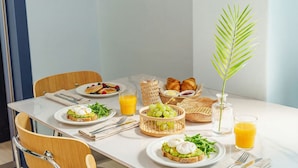 Uma mesa de café da manhã com pratos de comida, uma cesta de uvas, croissants e um vaso com uma planta.