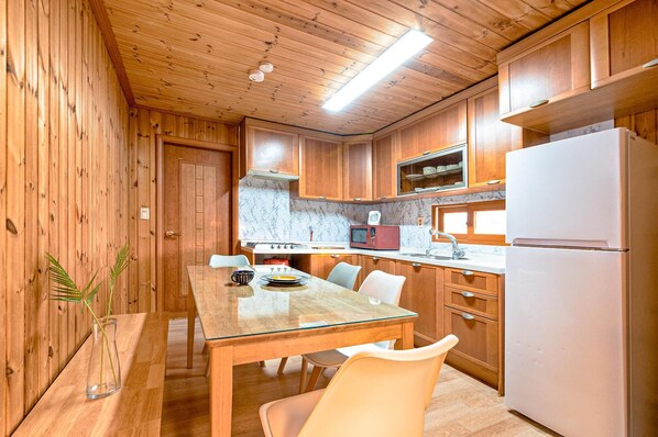 Interior de uma cabana de madeira com mesa de jantar, cadeiras, cozinha equipada com frigorífico, micro-ondas e fogão.